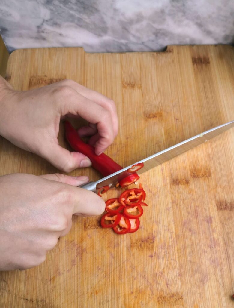 Hand chopping up red chilli with a large silver knife on a wooden board.